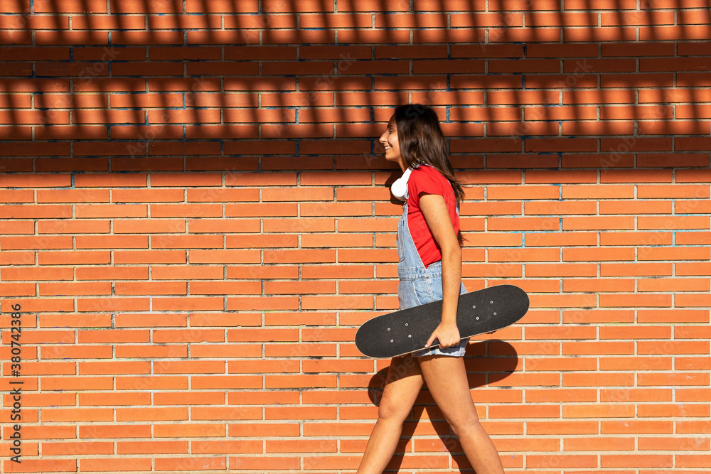 Side view of female millennial walking with skateboard near brick wall ...
