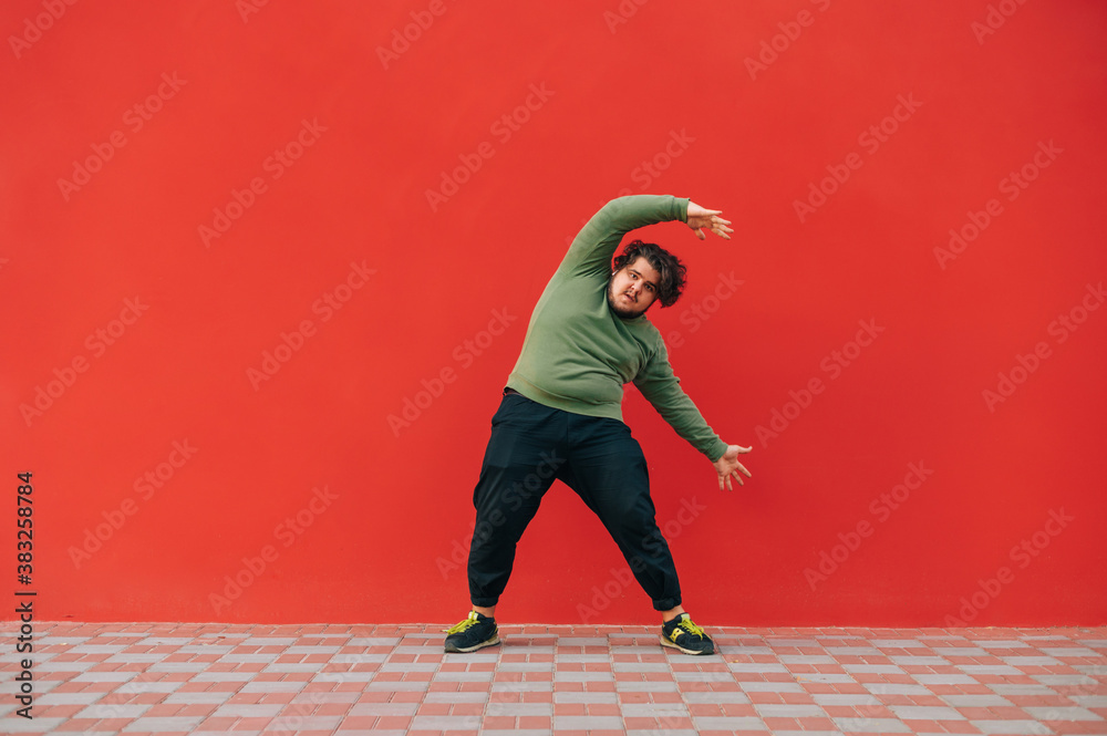 Funny fat man in casual clothes trains on a red wall, looks at the ...