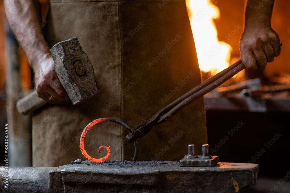 Old blacksmith is processing a hot metal object of a spiral shape on the anvil in the forge ...