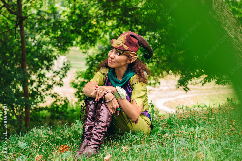 Sly look of a wood elf sitting on the grass in a magic forest. A woman ...