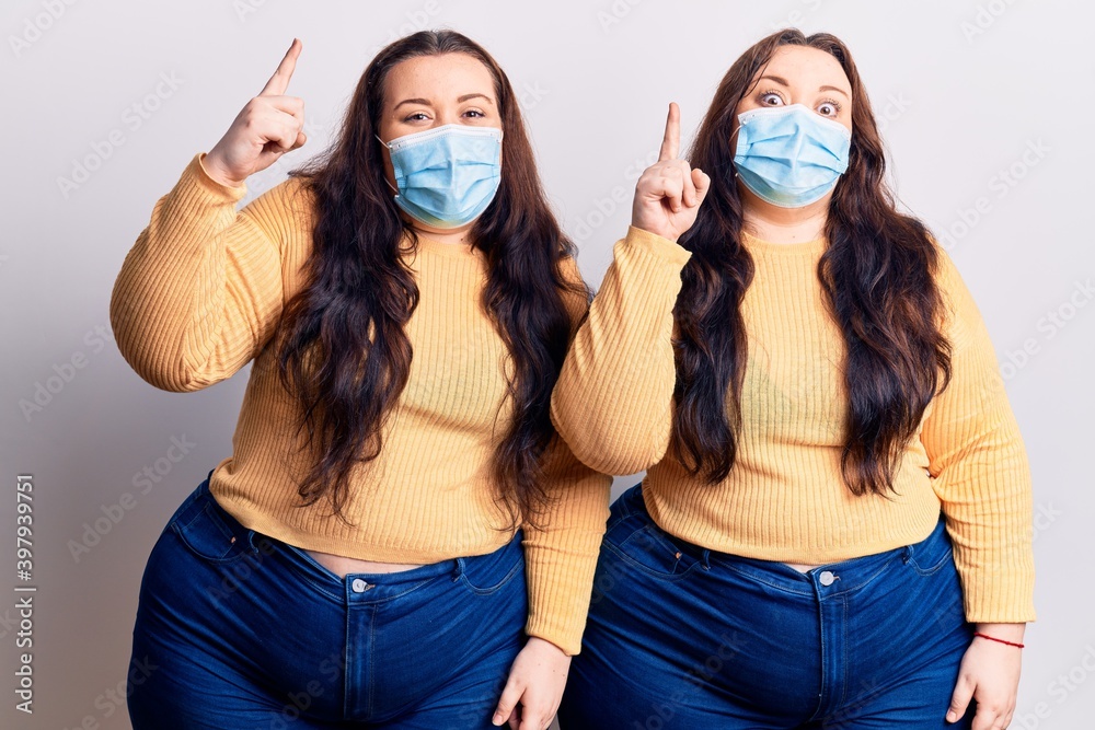 Young plus size twins wearing medical mask pointing finger up with ...