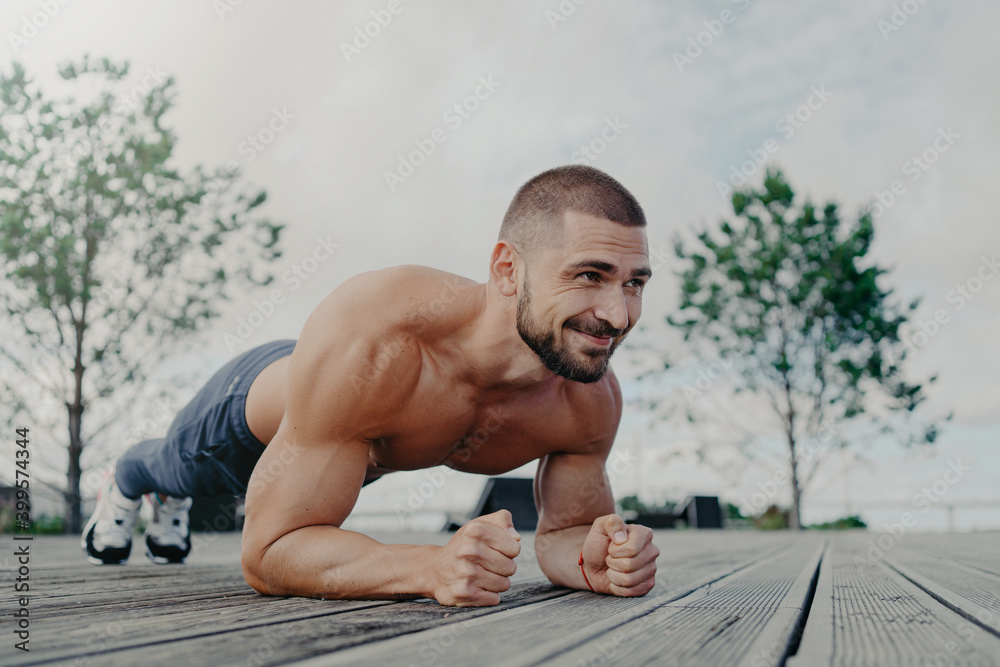 Shirtless determined bearded man with naked torso stands in plank pose and smiles gladfully ...