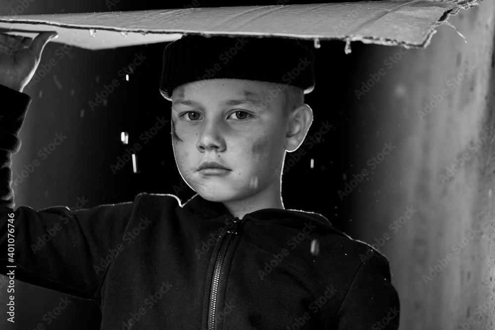 poor orphan boy shelters from the rain under cardboard, look at camera ...