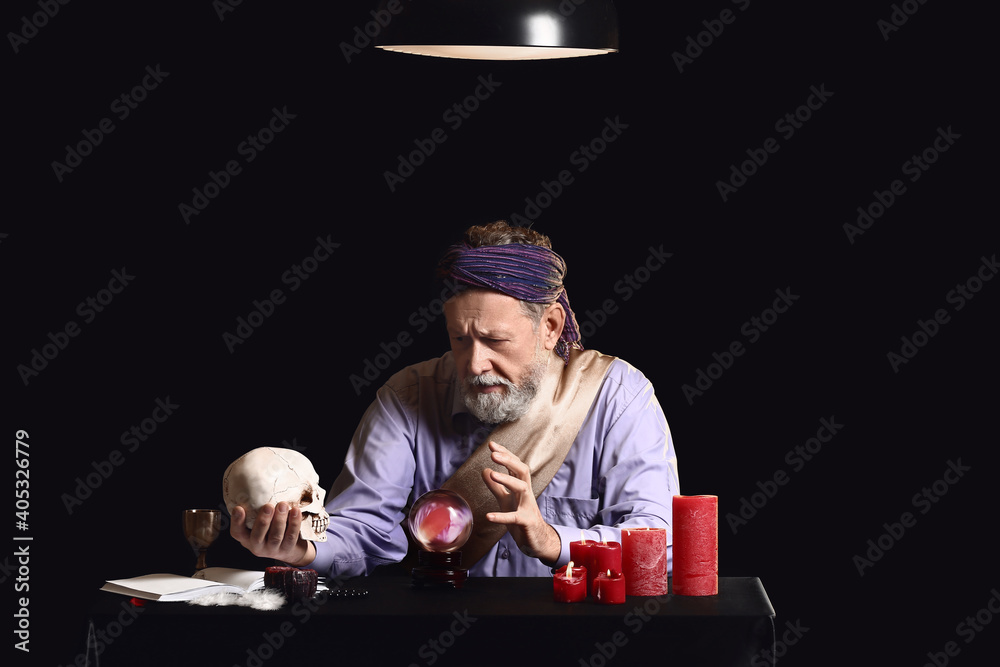 Male fortune teller with crystal ball and human skull at table on dark ...