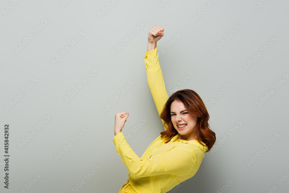 excited woman looking at camera and showing triumph gesture on grey ...