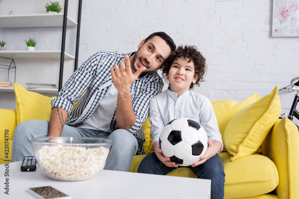 smiling muslim man pointing with hand near son while watching football ...