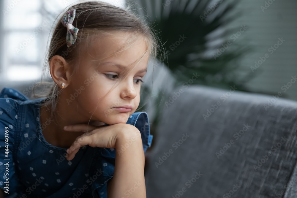 Close up of sad small Caucasian girl child sit on sofa look in distance ...