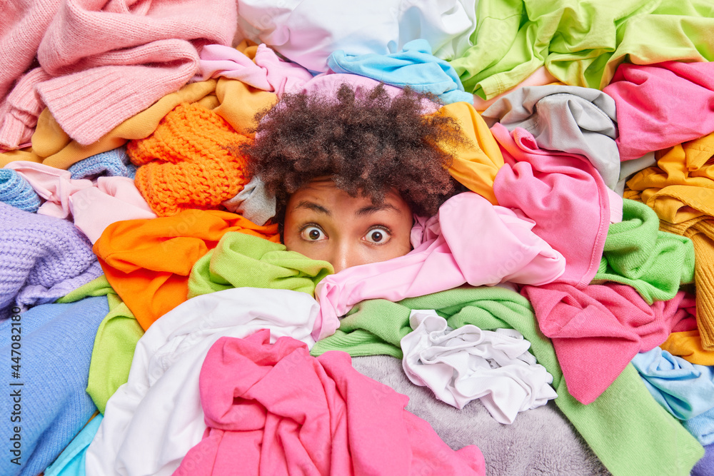 Surprised curly haired ethnic young woman stuck in pile of laundry does ...