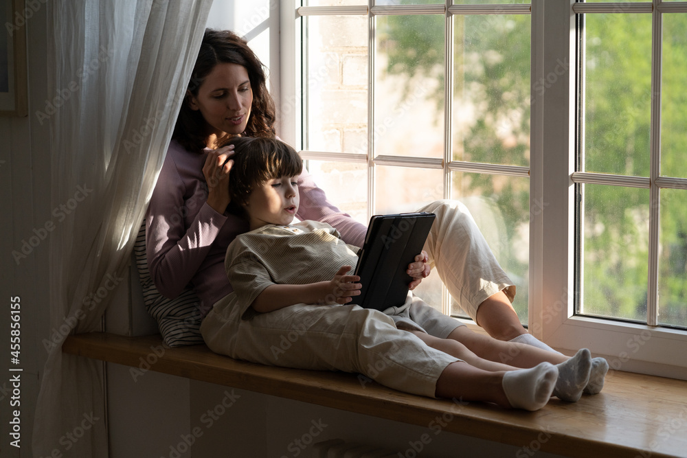 Young mom and kid boy sitting on windowsill look at digital tablet ...