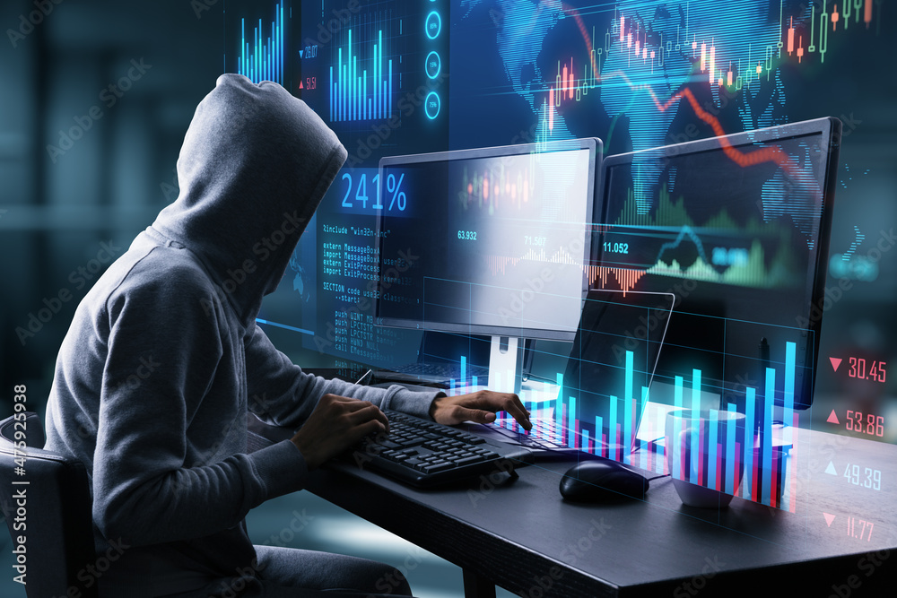 Hacker in hoodie using computers at desktop with glowing forex chart trading interface on blurry ...