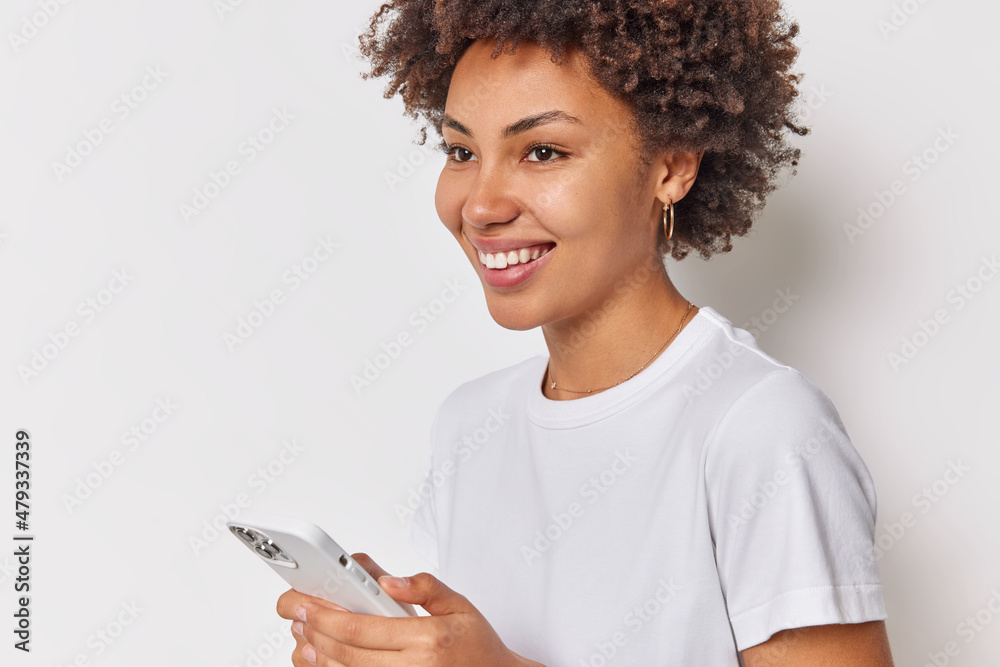 Happy positive young woman uses smartphone and chats online stands glad indoor receives pleasant ...