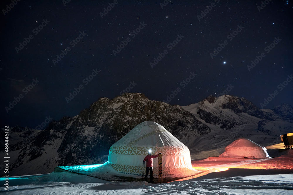 A man with a snowboard standing near a yurt at night in winter in the ...
