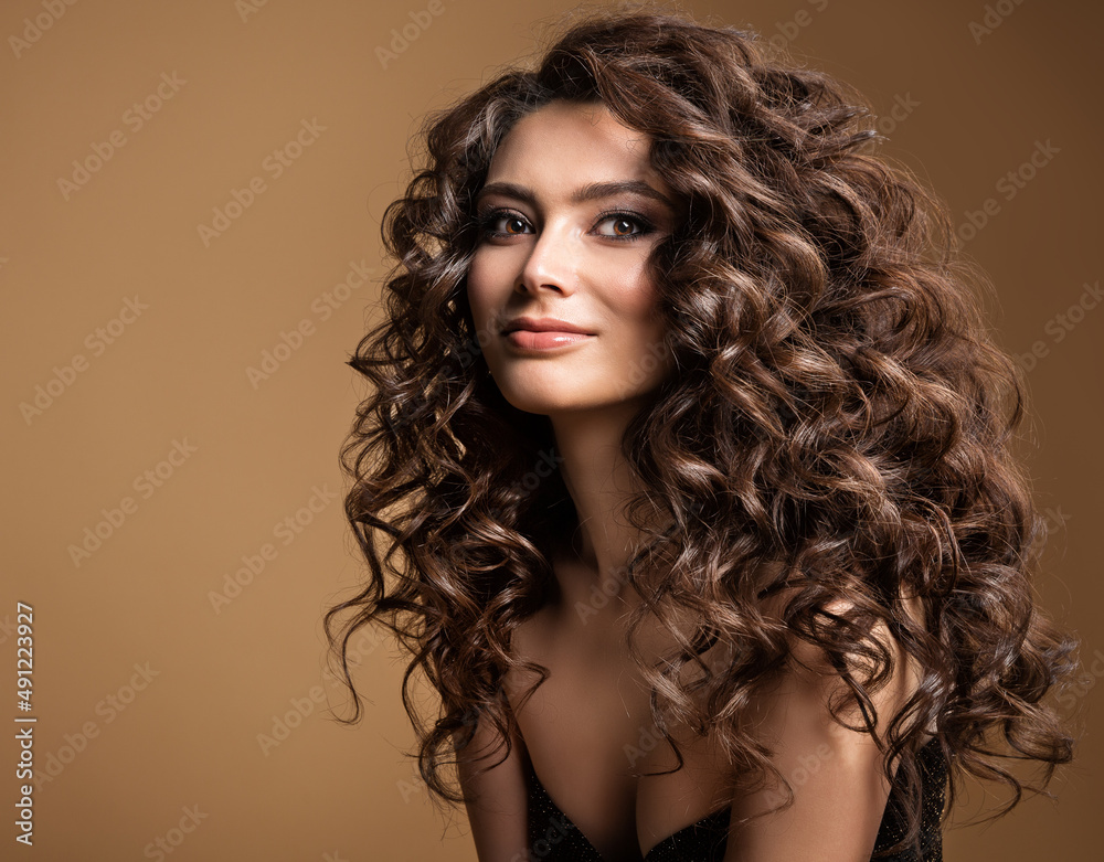 Curly Hair Model. Woman Wavy Long Hairstyle. Brunette Fashion Girl with Volume Hairdo and Natural Make up over Beige Background Adobe Stock