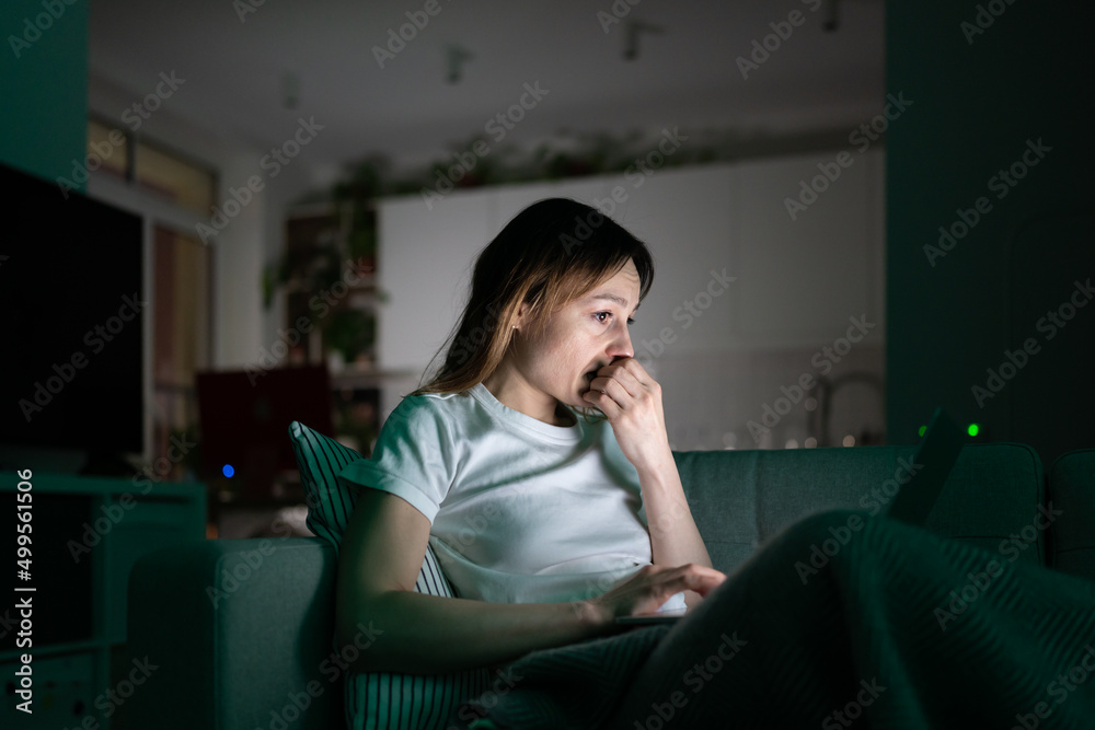 Young anxious woman lying on sofa staring at laptop screen at night ...