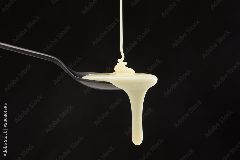 Sweetened condensed milk pouring down from spoon on a black background ...