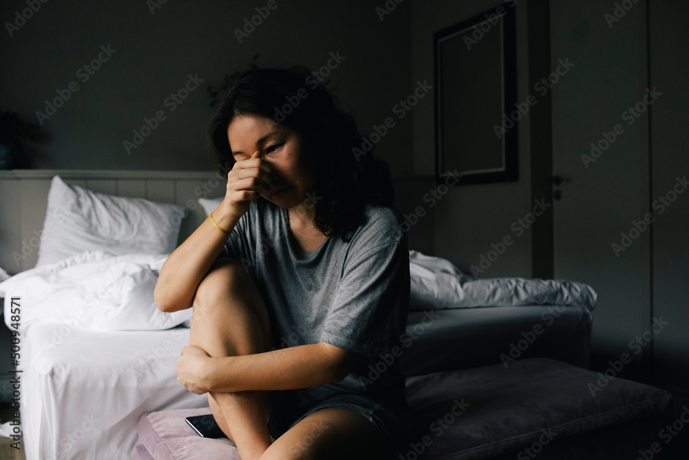 Sad woman crying while sitting on bed end bench in bedroom at home ...