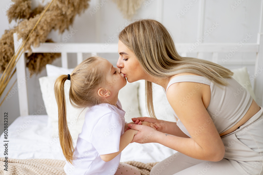mom and daughter play, hug and kiss at home on the bed, lifestyle, tender relationship of a ...