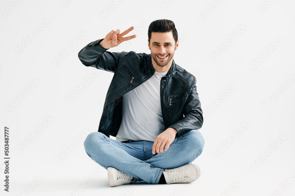 Positive cheerful young man doing victory sign sitting with legs ...
