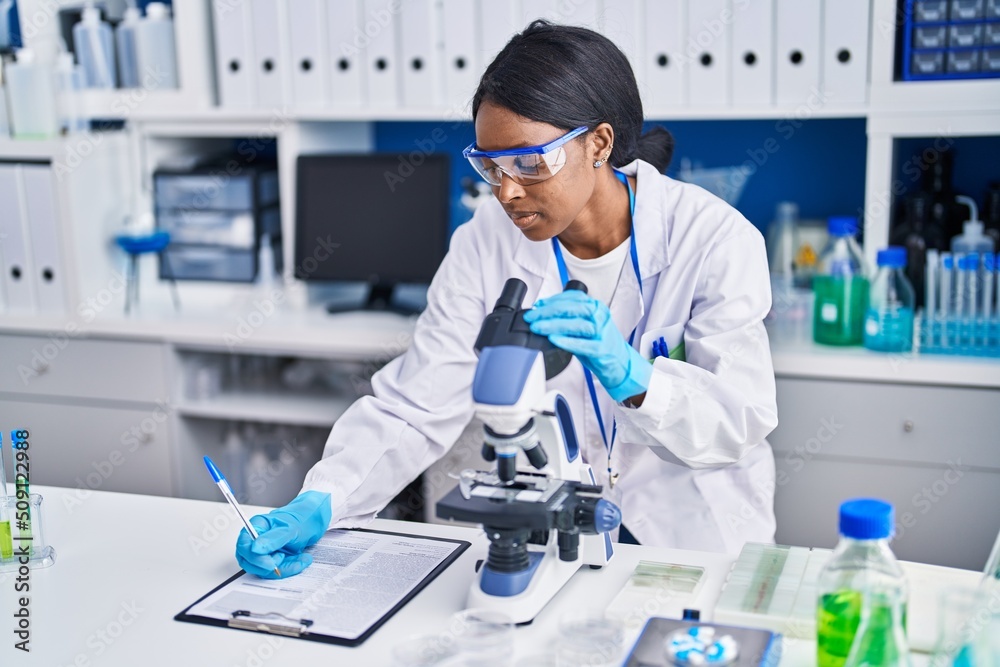 Young african american woman scientist using microscope write on document at laboratory Stock ...