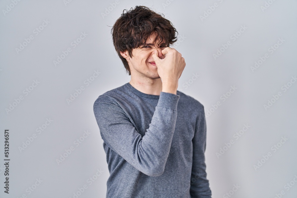 Young man standing over isolated background smelling something stinky ...