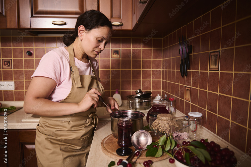 Charming housewife in apron, stands by kitchen countertop and pours ...