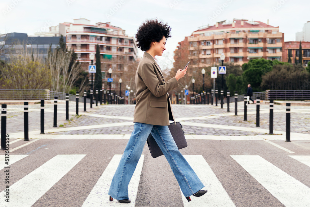 smiling latin business woman walking on a crosswalk using her smart ...