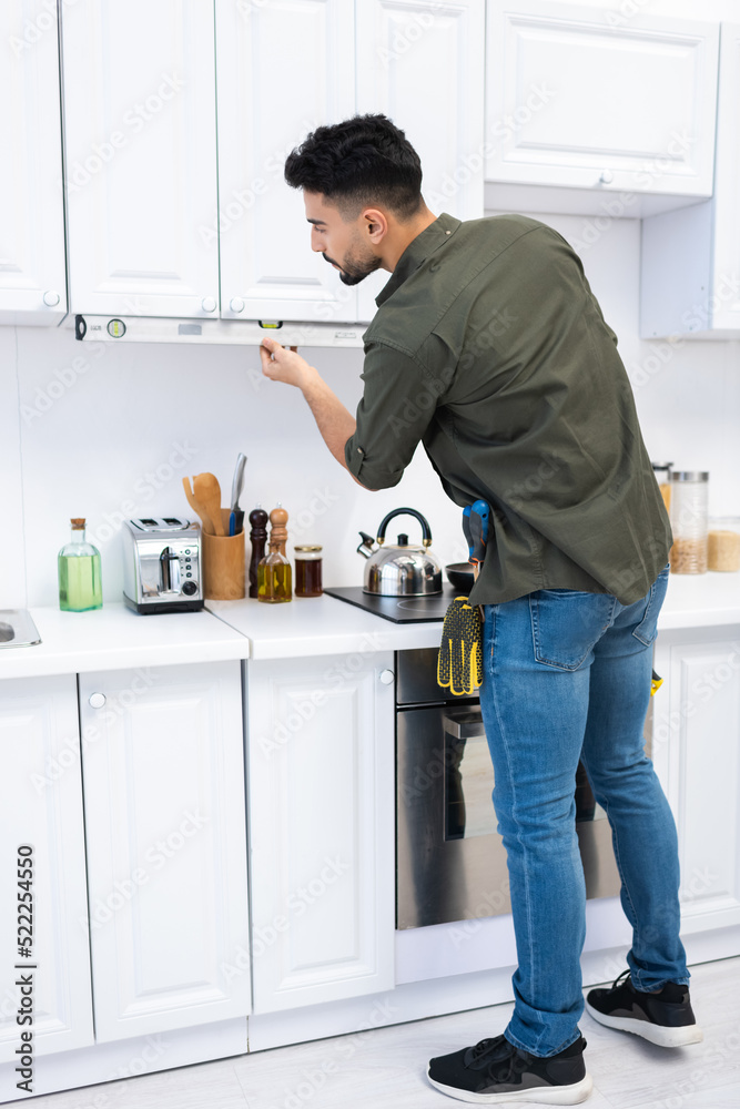 Side view of arabian man with spirit level measuring kitchen cupboard ...