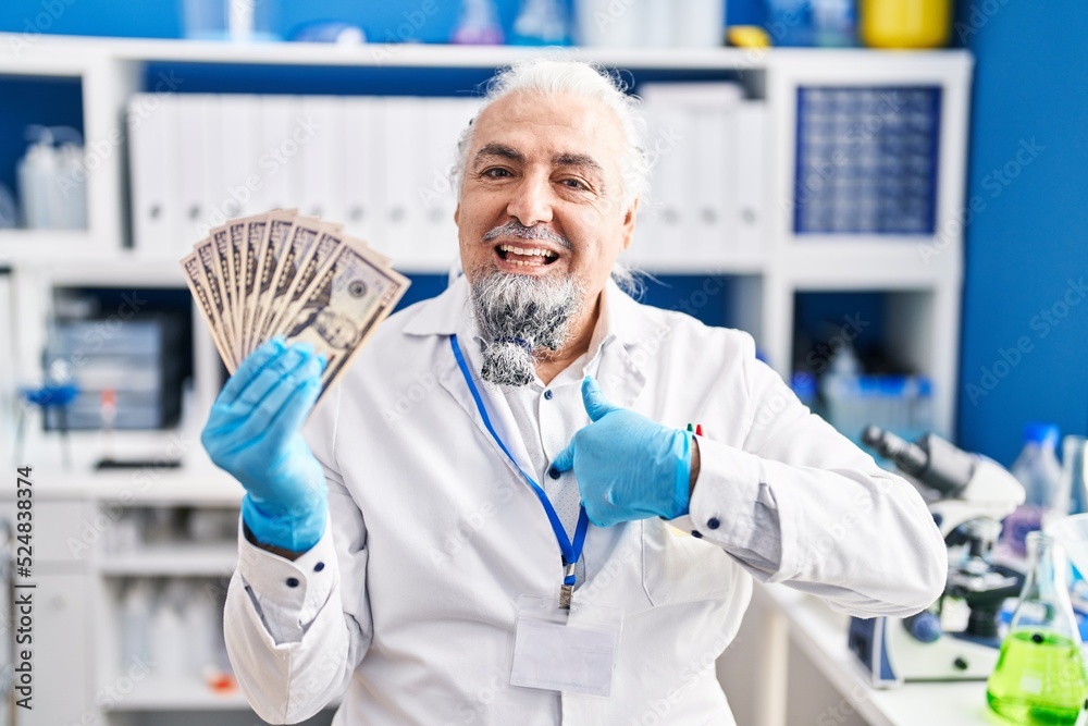 Middle age man with grey hair working at scientist laboratory holding money pointing finger to ...