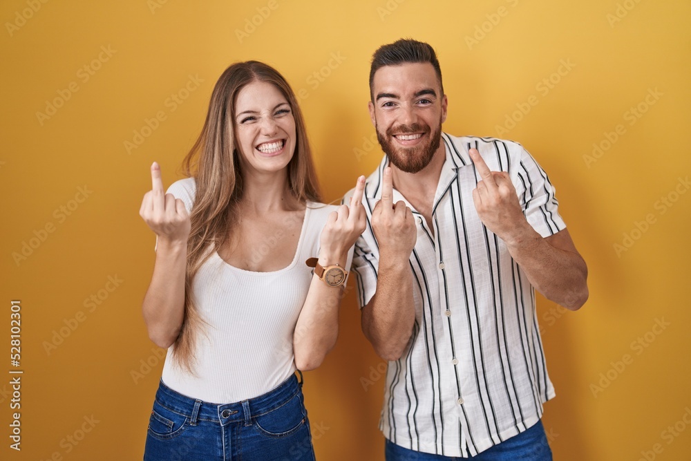 Young couple standing over yellow background showing middle finger ...