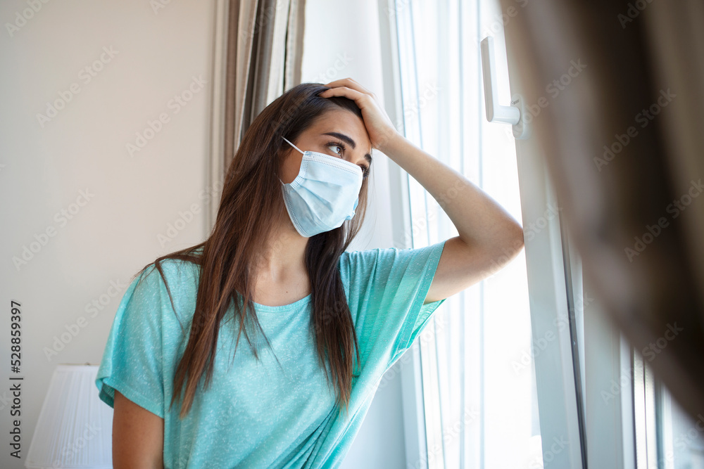 Young woman wearing protective face mask looking outside window with ...