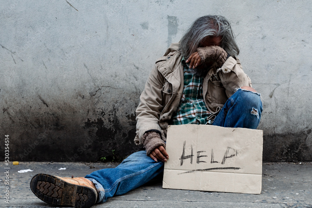 homeless, long-haired Asian man sits hopelessly leaning against a wall ...
