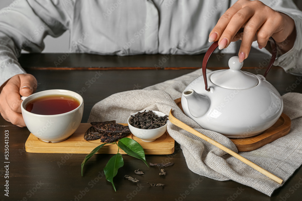 Female hands with cup of hot puer tea, teapot and dry leaves at dark ...