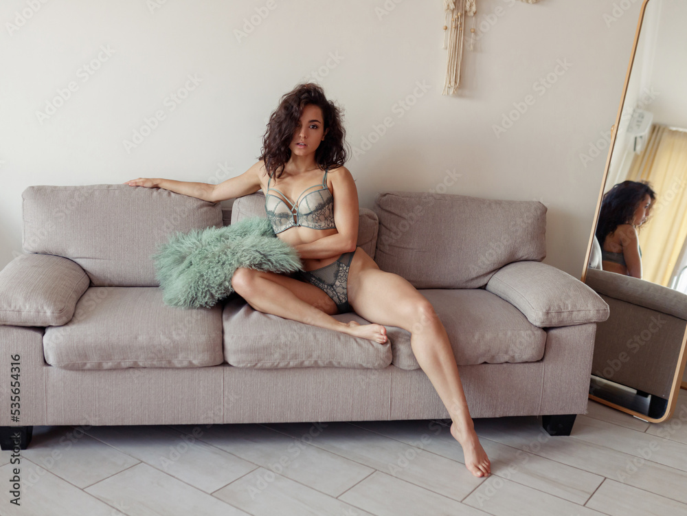 Young beautiful woman in sexy lingerie sitting on a sofa in modern room at home looking at ...