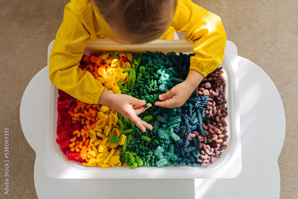 Child playing with sensory bin with dried pasta in rainbow colors. Dyed ...