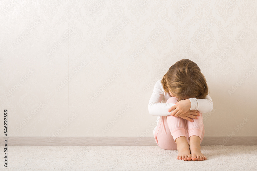 Sad alone little girl crying and sitting on carpet at home room. Child hugging knees with hands ...