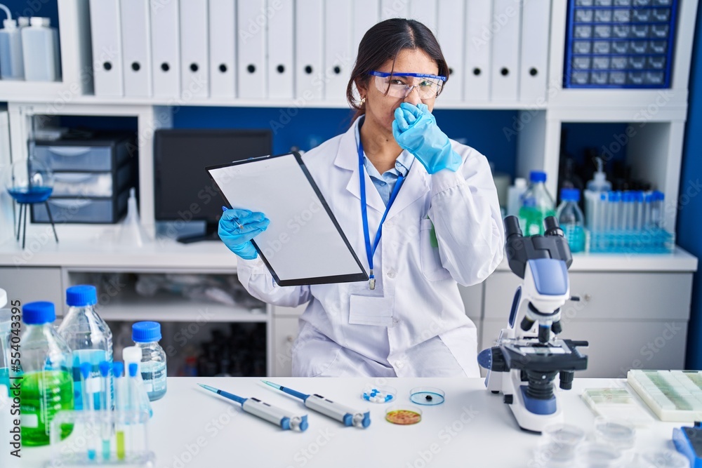 Hispanic young woman working at scientist laboratory smelling something ...