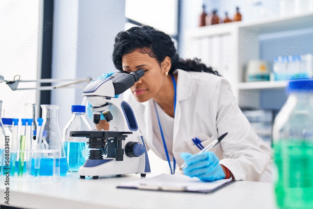 African american woman wearing scientist uniform using microscope write on document at ...