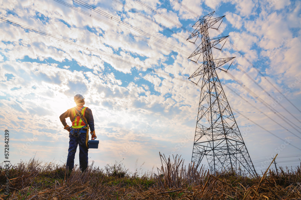Technician stands carrying a toolbox to a high-voltage pylon wearing an ...