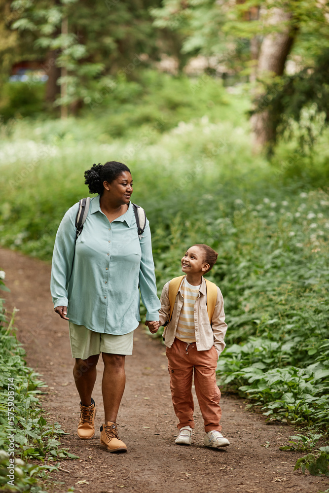 Vertical portrait of black mother and daughter walking on nature trail together holding hands ...