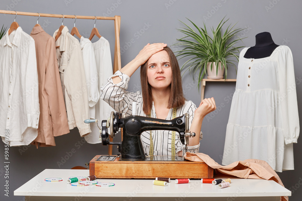 Sad upset woman sewer posing at her workplace sewing clothing, showing ...