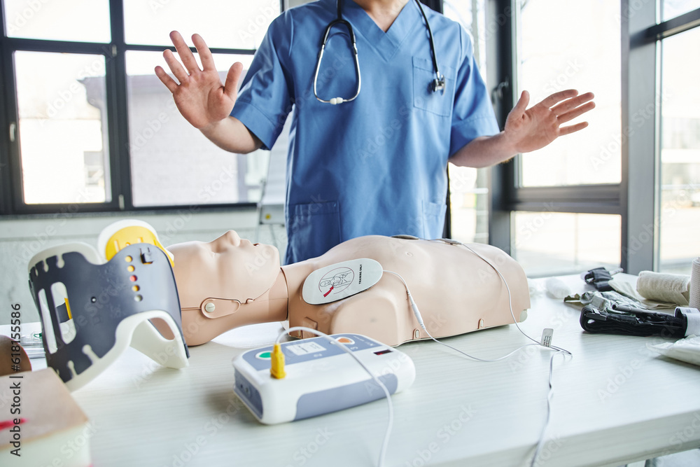 cropped view of healthcare worker in blue uniform gesturing near CPR manikin with defibrillator ...