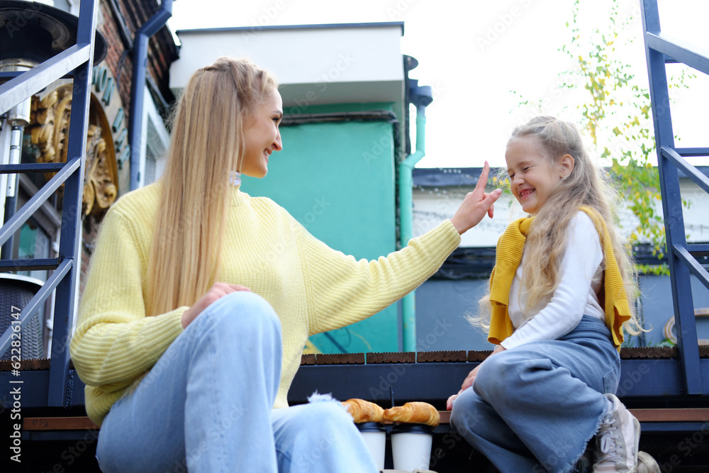 Mom and daughter are sitting on the steps of the city cafe and fooling ...