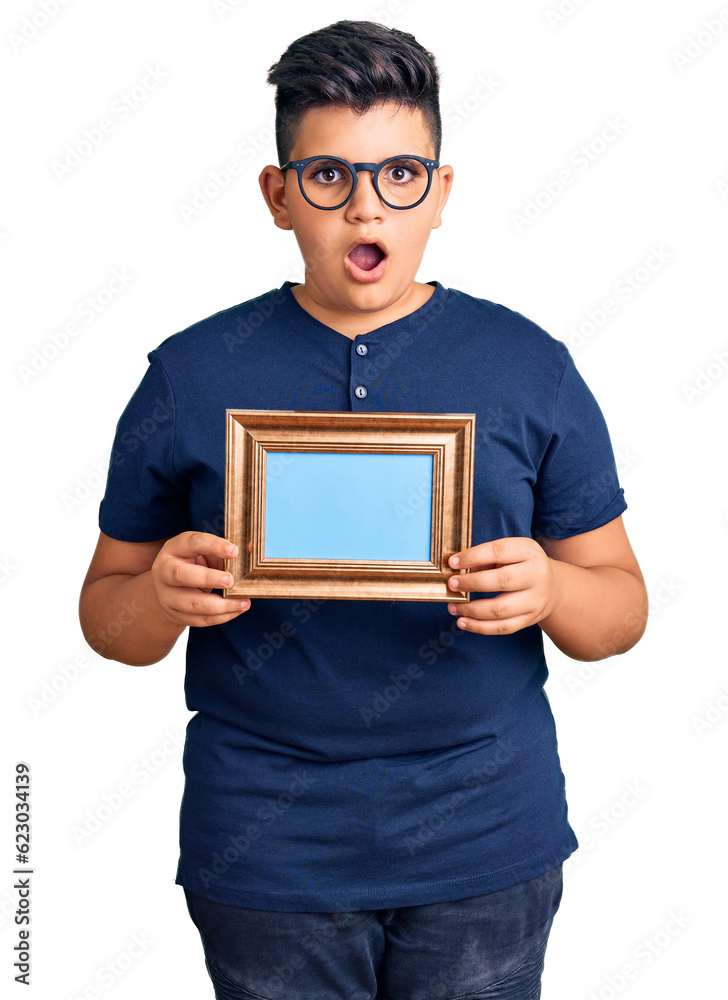 Little boy kid holding empty frame scared and amazed with open mouth for surprise, disbelief ...