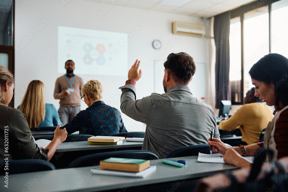 Back view of older student raising his hand to answer teacher's ...