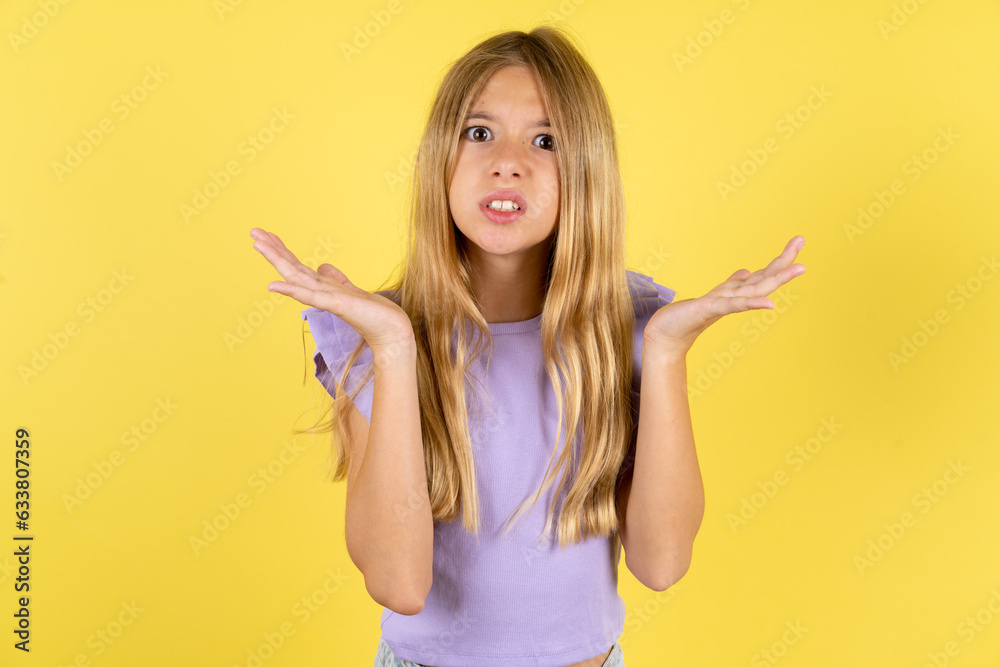 Indignant blonde kid girl wearing violet T-shirt over yellow studio ...
