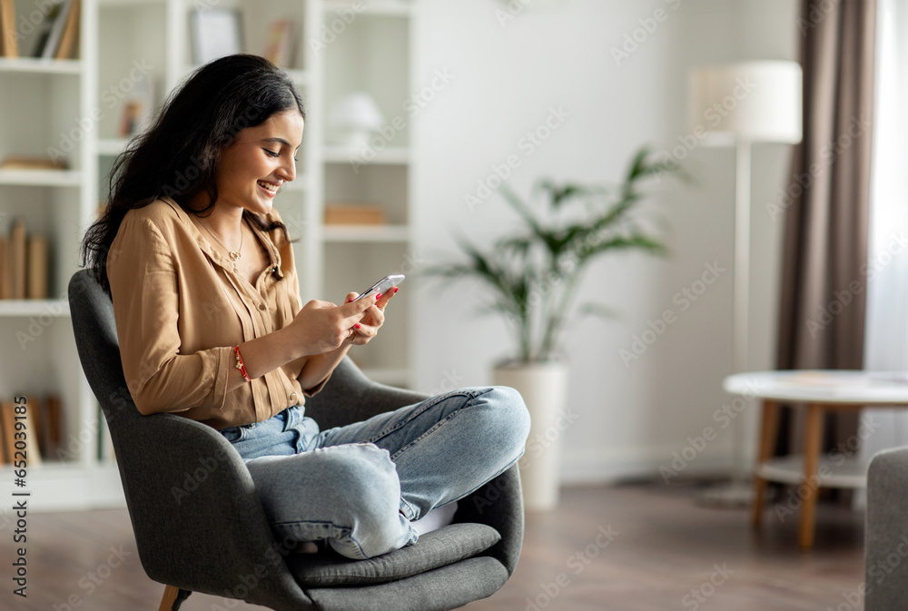 Happy indian woman using cellphone, sitting in armchair, browsing ...