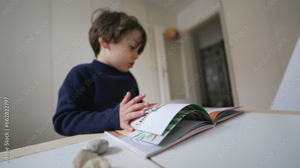 Child turning book pages looking at images and text. Little boy reading ...
