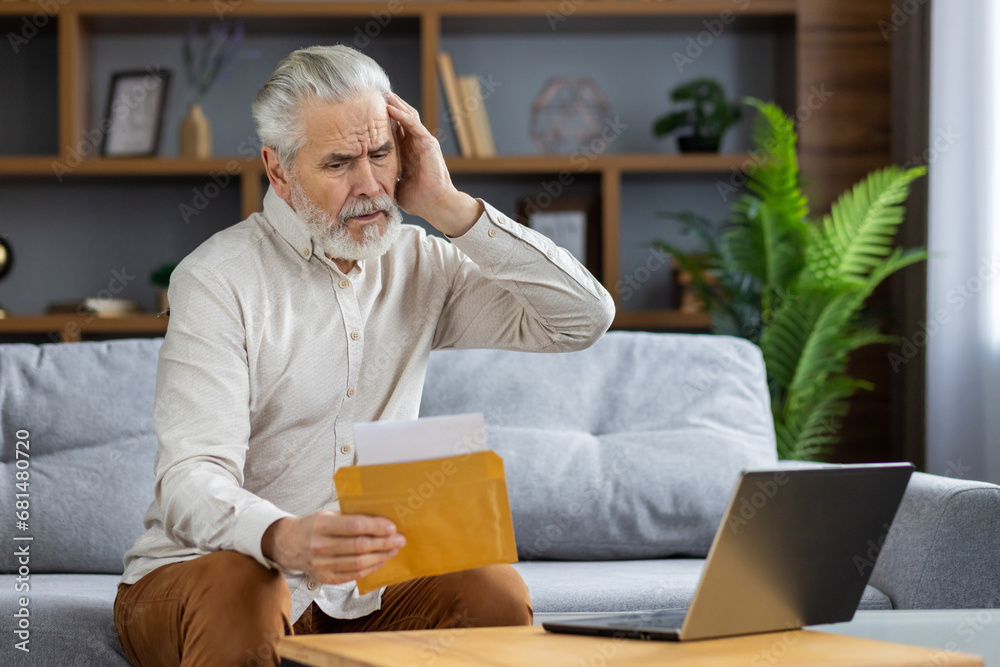 Upset senior gray-haired man sitting at home on sofa in front of laptop ...