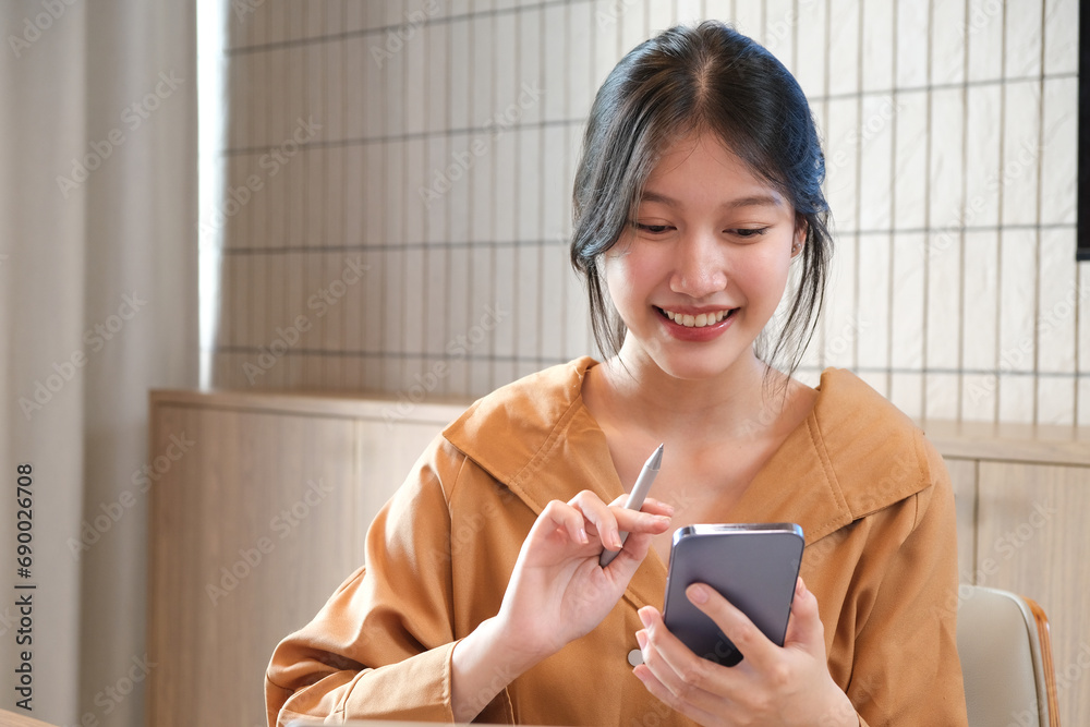 Smiling and charming Asian woman sit at her desk with a smartphone ...