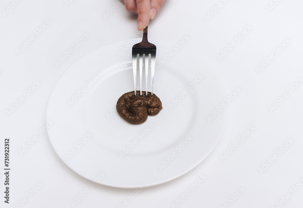 A hand with a fork is touching artificial feces lying on a white plate ...